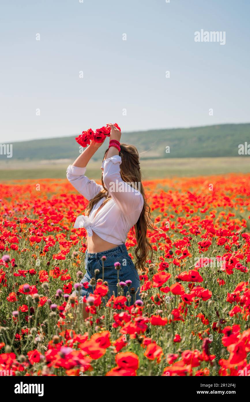 Glückliche Frau in einem Mohnfeld in einem weißen Hemd und Jeansrock mit einem Mohnkranz auf dem ...