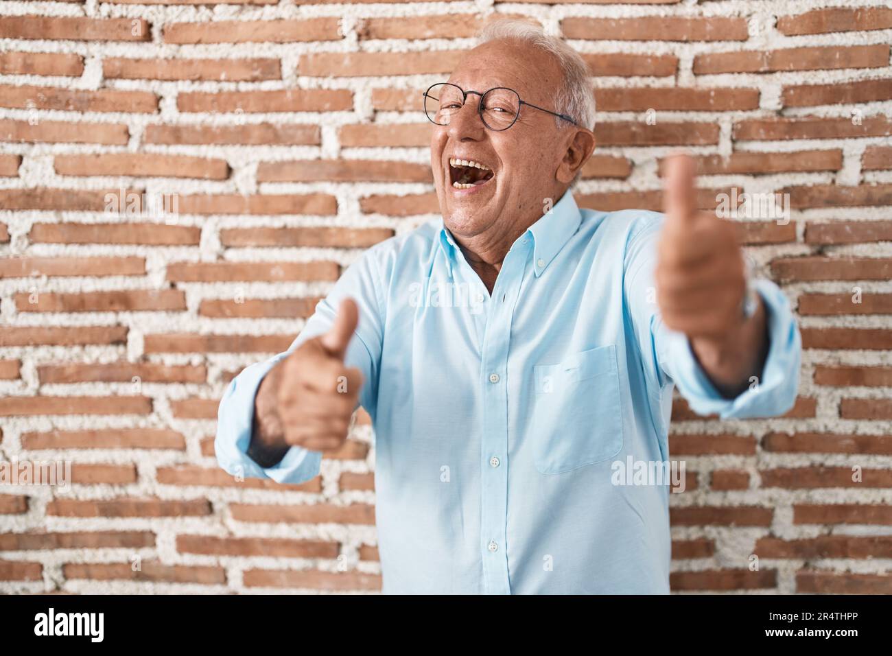Älterer Mann mit grauen Haaren, der über einer Ziegelwand steht und eine positive Geste mit der ...