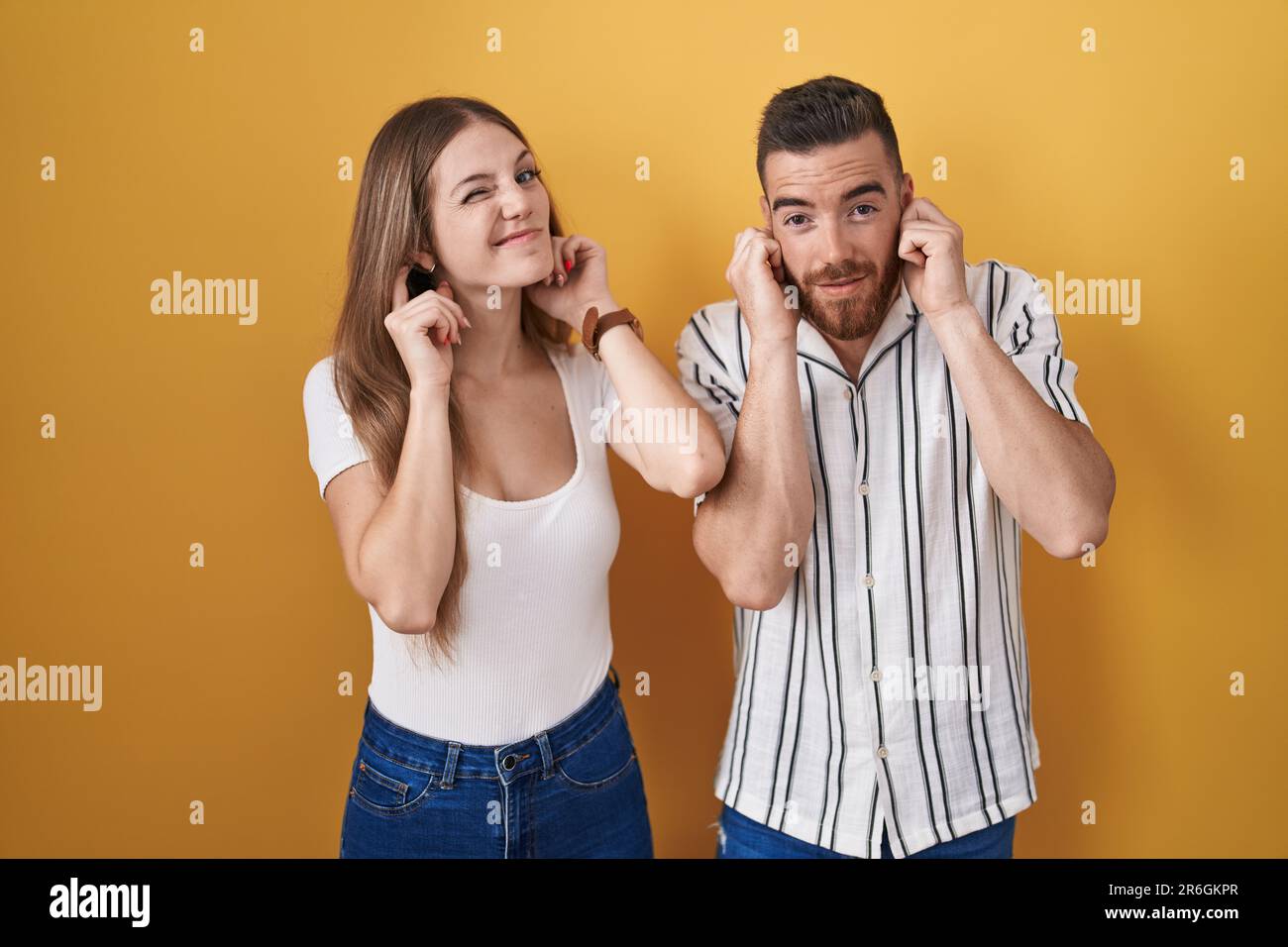Ein junges Paar, das auf gelbem Hintergrund steht und die Ohren mit ...