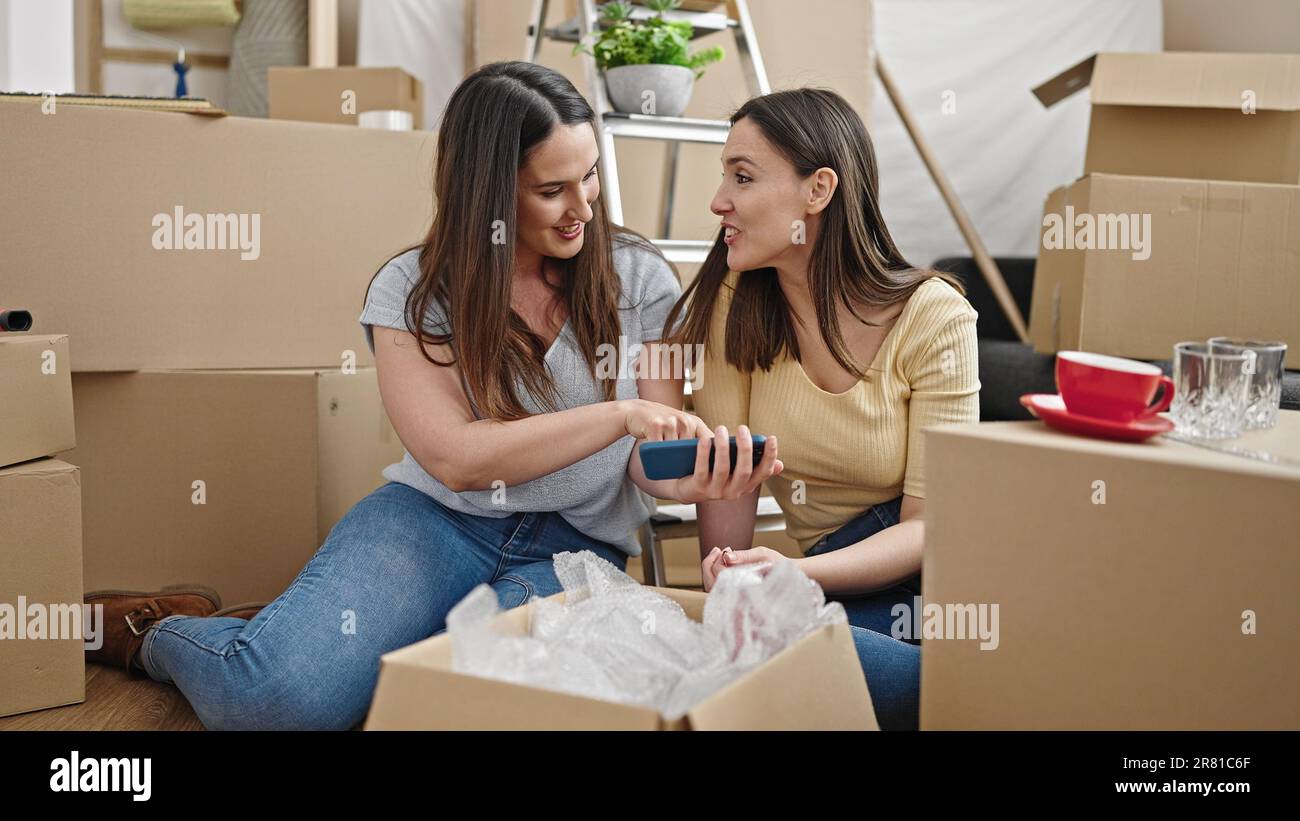 Zwei Frauen, die ihr Smartphone benutzen, sitzen in ihrem neuen Zuhause ...