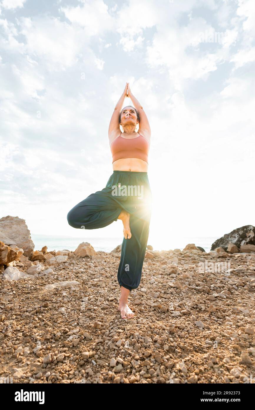 Eine Frau, die Yoga im Baum praktiziert und am Strand vor dem Himmel ...