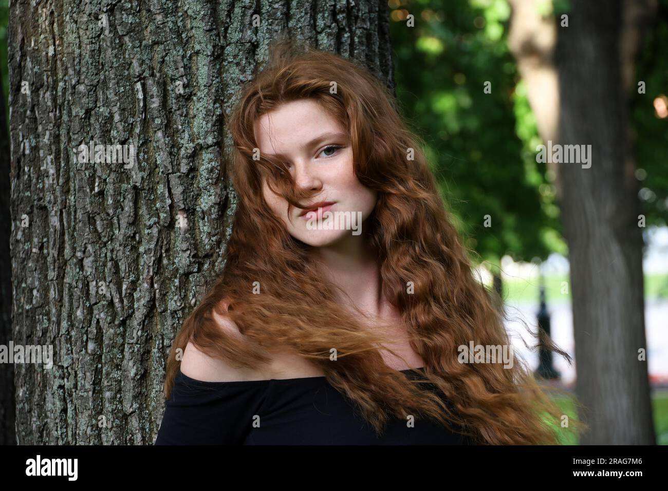 Ein sinnliches Mädchen mit langen roten Haaren und Sommersprossen steht neben dem Baum im ...