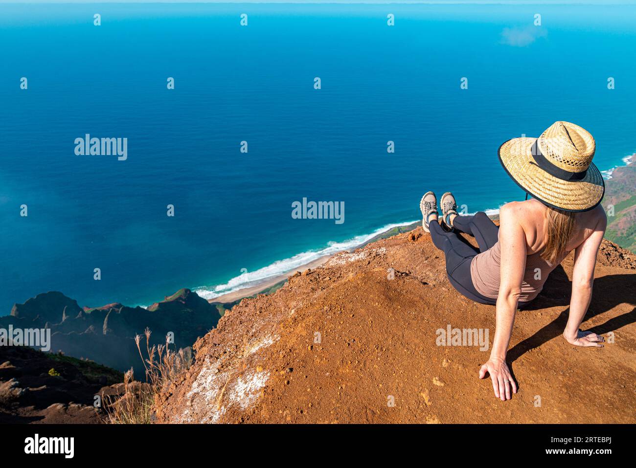 Blick von hinten auf eine Frau mit Strohhut, die auf einer Bergklippe ...