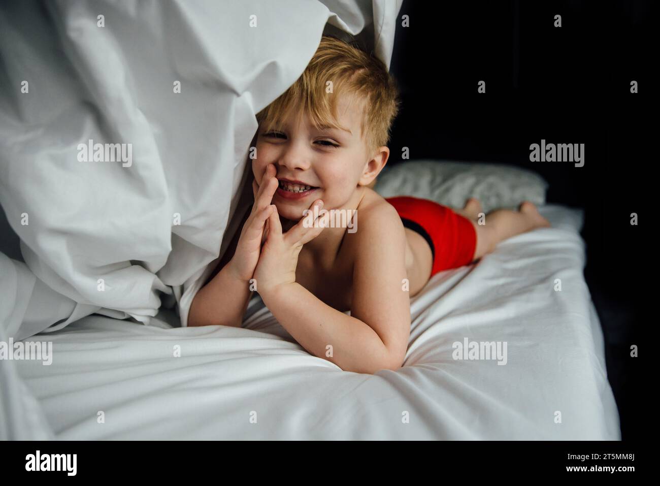 Der kleine Junge liegt in Shorts auf dem Bett und lächelt dabei in die Kamera Stockfotografie ...
