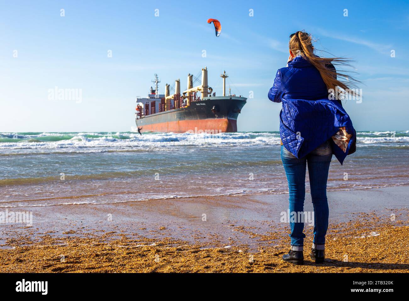 Eine Frau filmt auf ihrem Handy ein großes Frachtschiff an der Küste ...