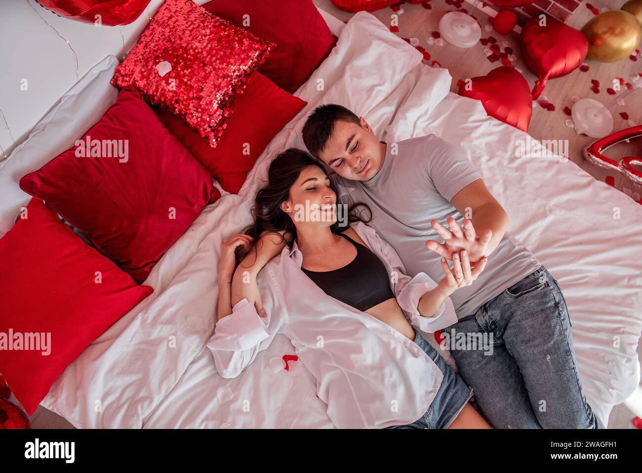Blick von oben auf ein liebevolles Paar in legeren Kleidern auf weißem Bett mit roten ...