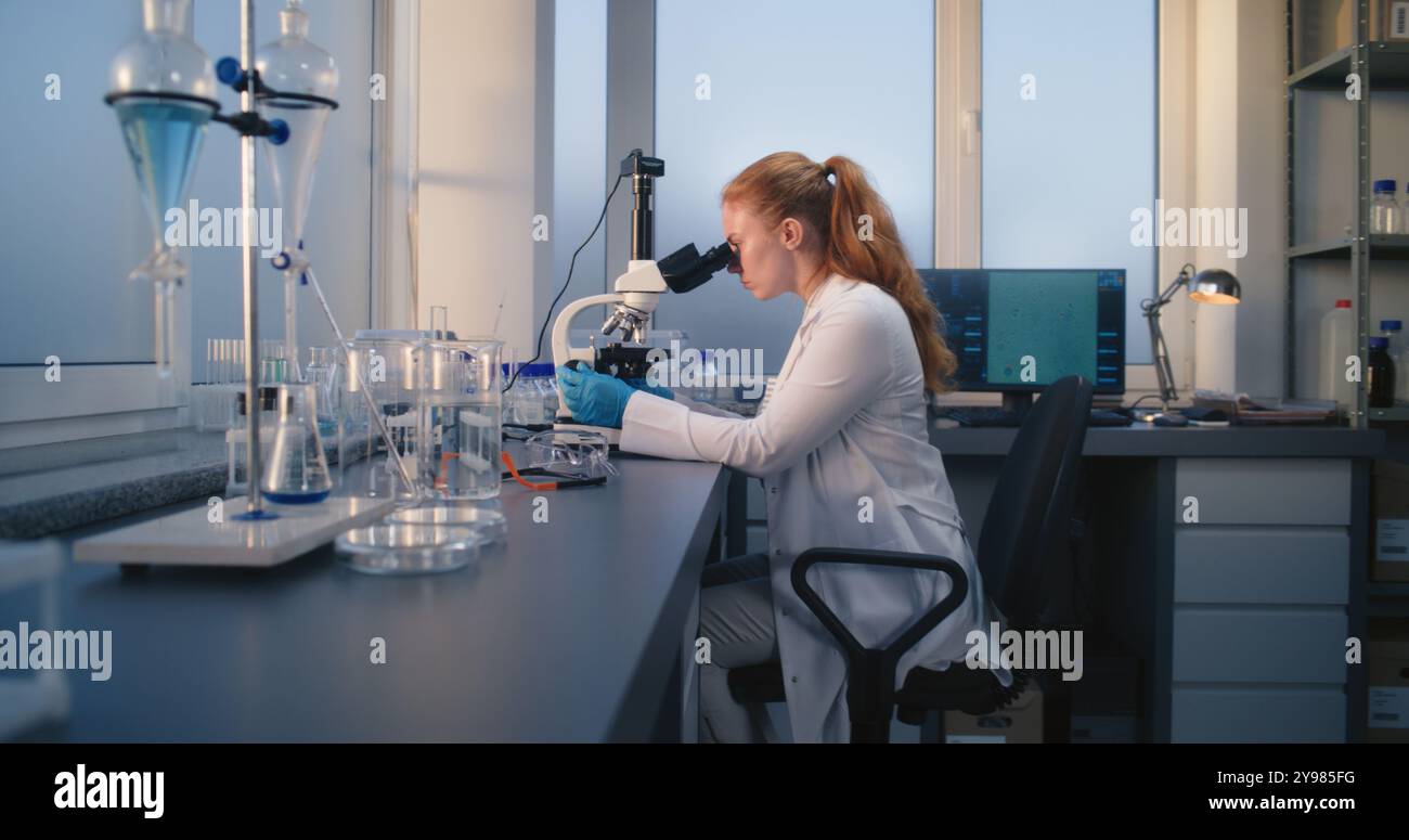 Junge Wissenschaftlerin sitzt im Stuhl, schaut unter dem Mikroskop und ...