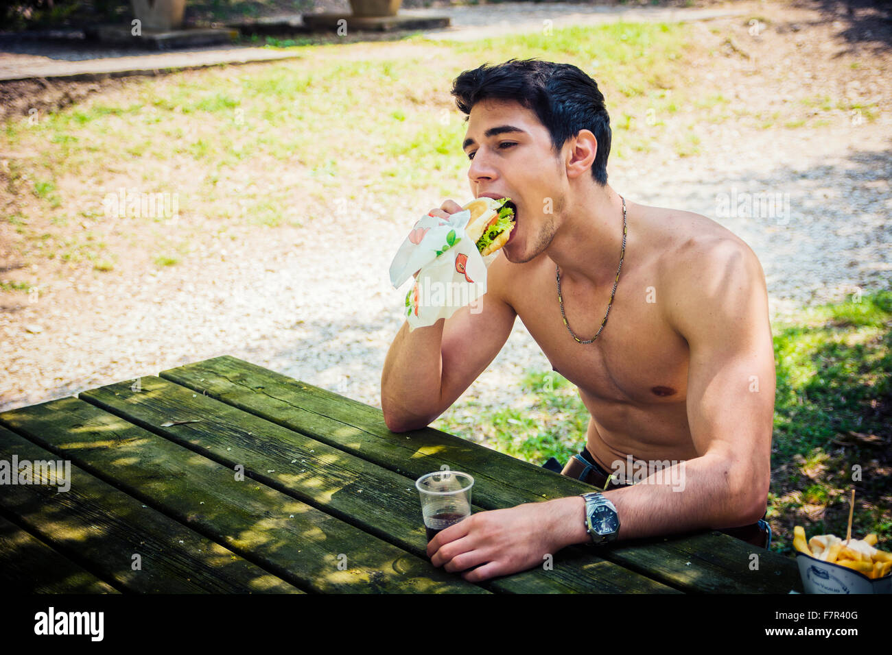 Porträt des jungen muskulösen Mann sitzen im Park am Picknicktisch essen Fast-Food - Burger ...