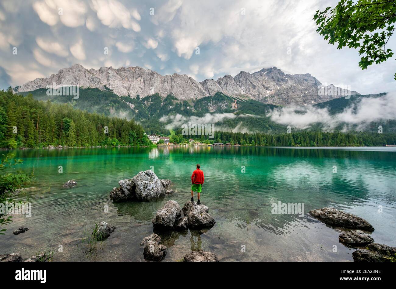 Jeune homme debout sur un rocher sur le rivage, lac Eibsee en face du massif de Zugspitze avec ...