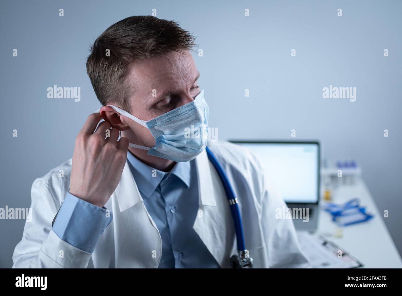 Un jeune médecin exténué en blouse de laboratoire enlève un masque et ...