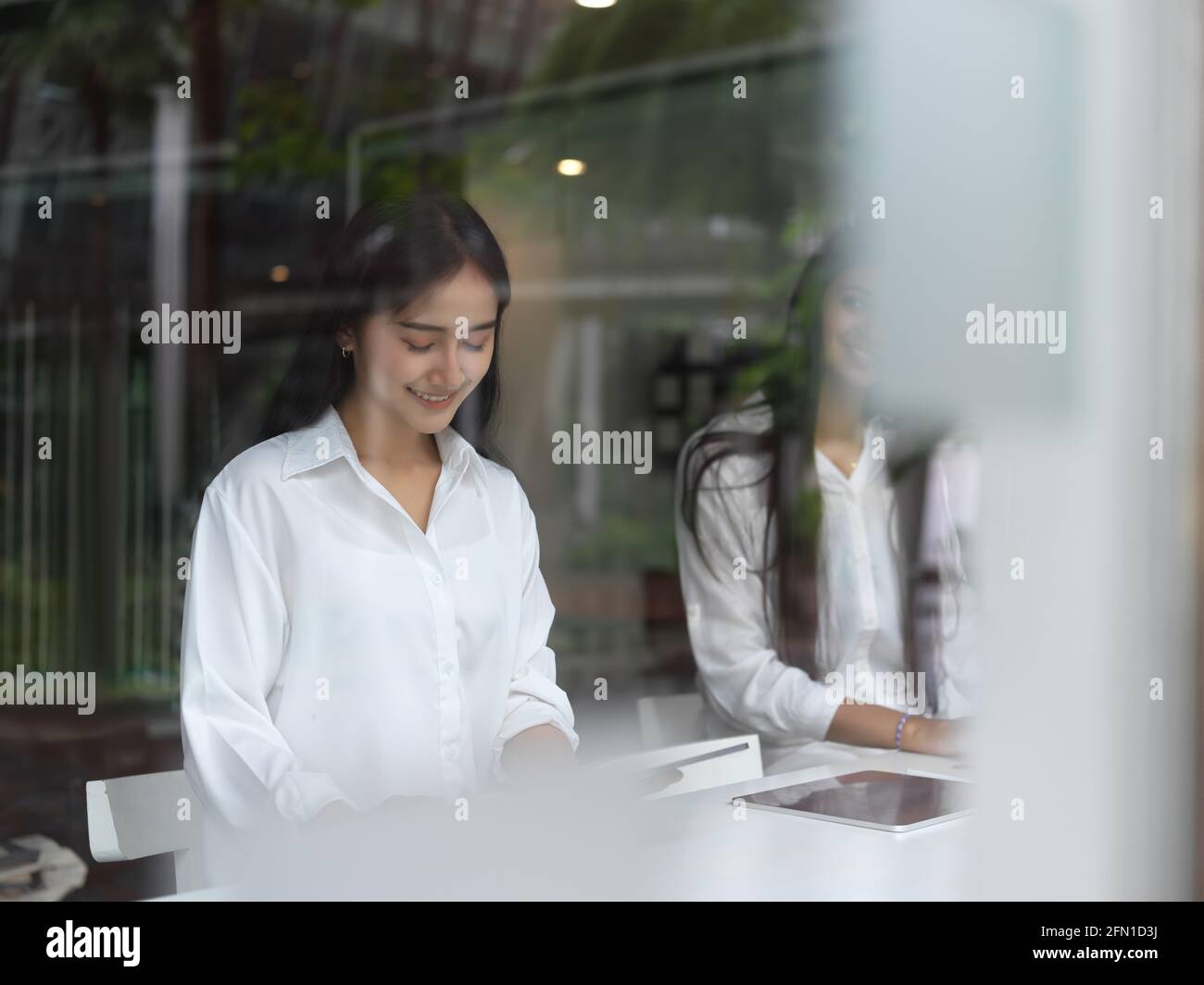 Vue à travers la fenêtre en verre de la femme se concentrant sur la tablette numérique tout en ...