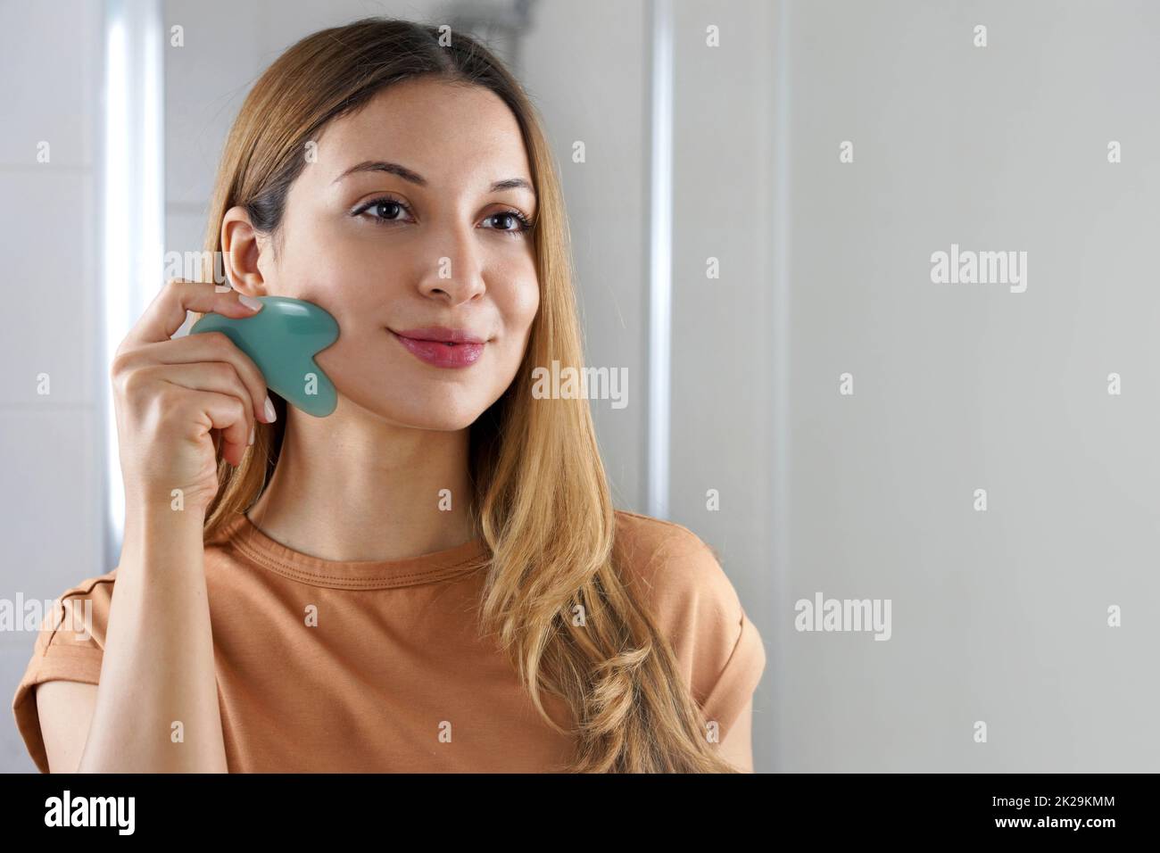 Portrait de la belle jeune femme regarde dans le miroir à l'aide de la carte de gua sha jade ...