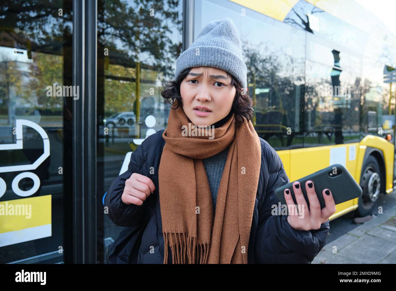 Portrait d'une fille asiatique confuse, debout à l'arrêt de bus, tenant ...
