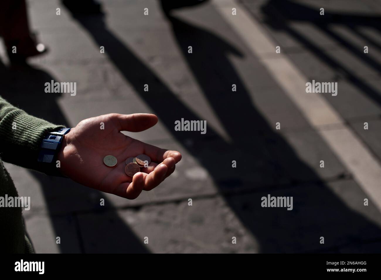 A man begging on a street, at left, casts a shadow as people walk past ...