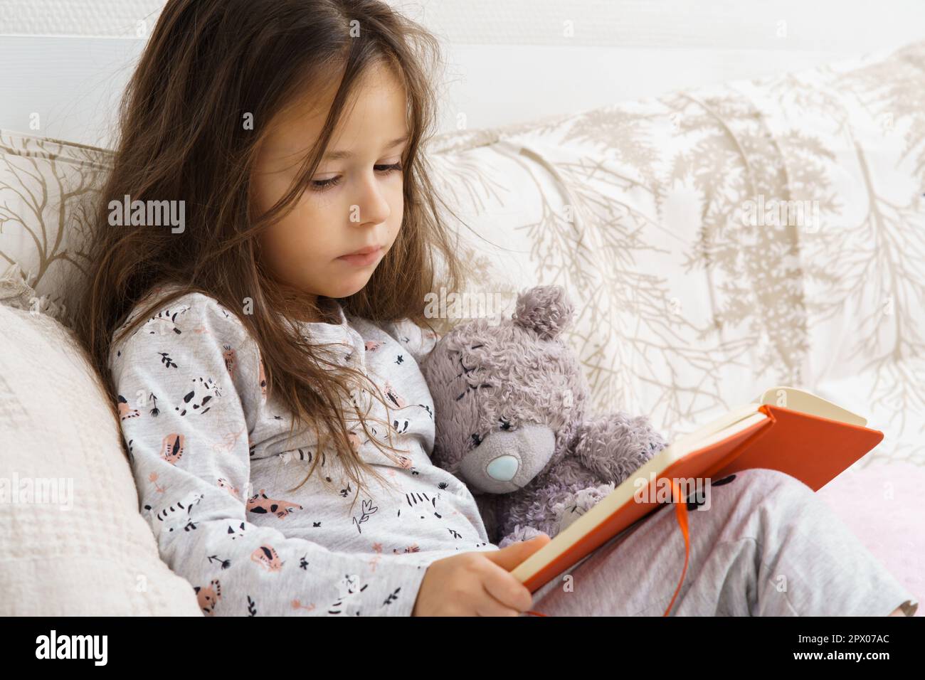 Petite fille malheureuse en vêtements de nuit assis sur le canapé et livre de lecture. Portrait ...