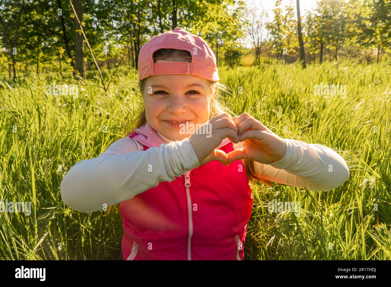 Une jeune fille gaie est assise dans l'herbe verte épaisse, représentant un signe de coeur avec ...
