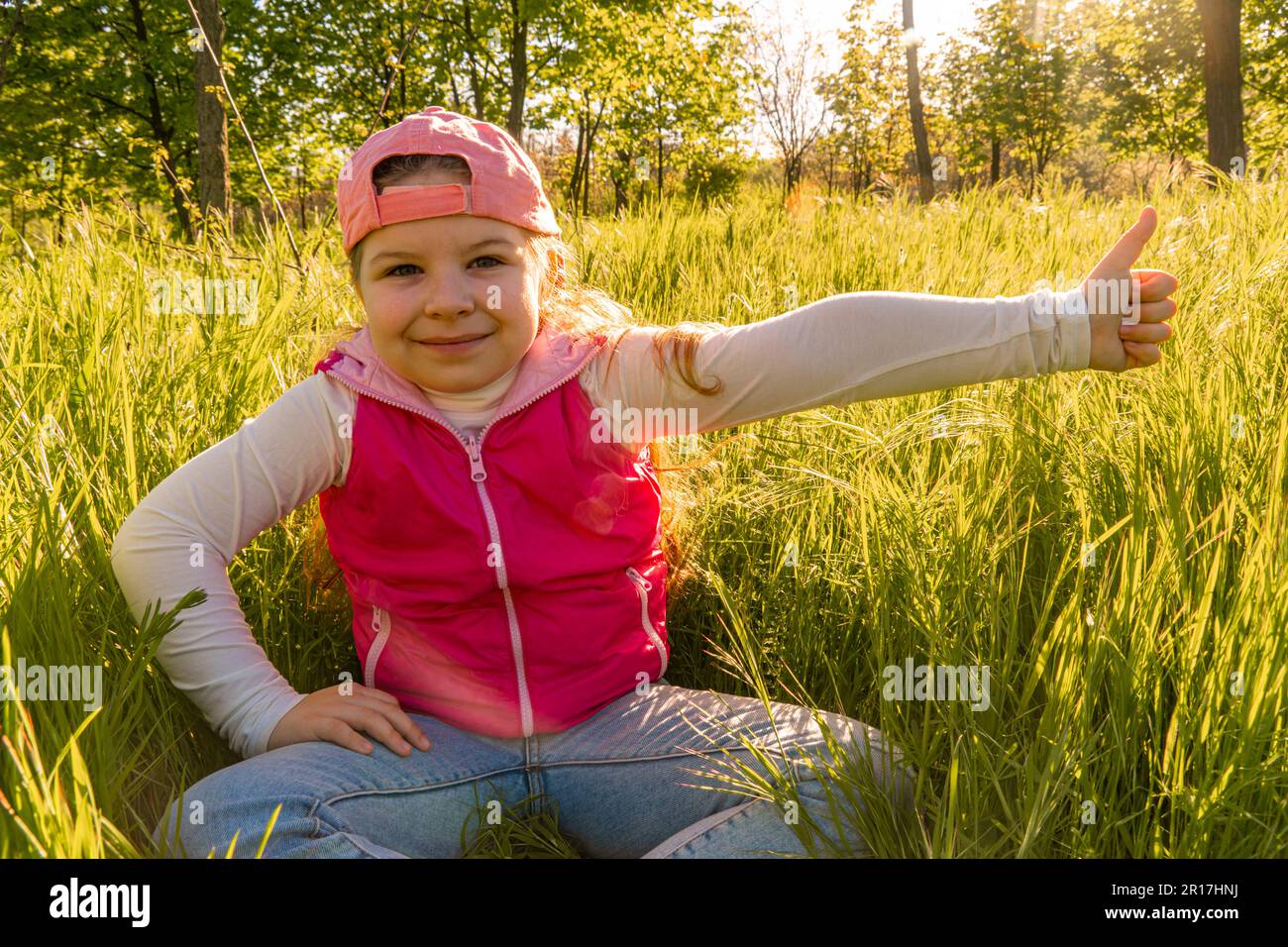 Une jeune fille gaie est assise dans l'herbe verte épaisse et fait un geste de main avec un ...