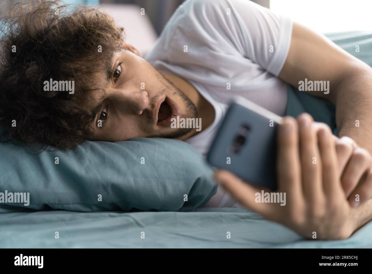 Un jeune homme choqué au lit regardant l'écran du smartphone à la ...