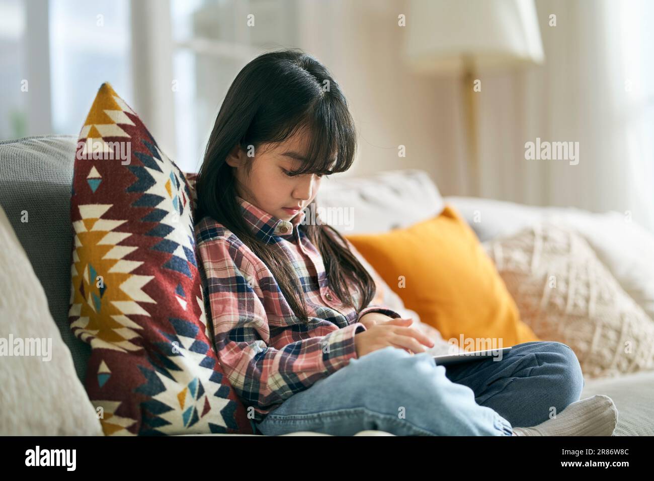 petite fille asiatique de sept ans assise sur un canapé de famille à la maison à l'aide d'un ...