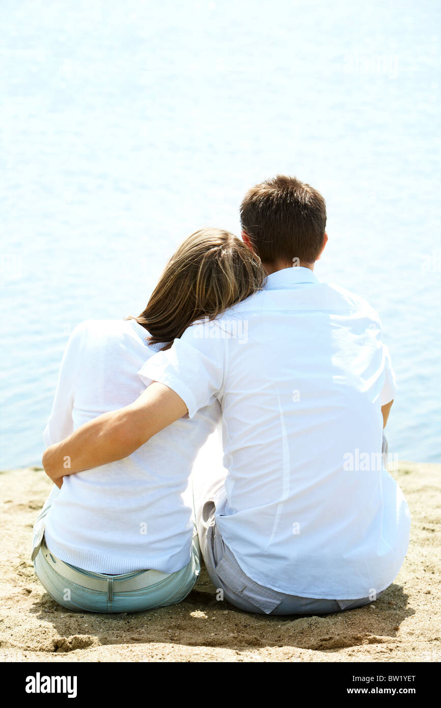 Dos de serene couple assis sur la rive de sable en face de l'eau bleue ...