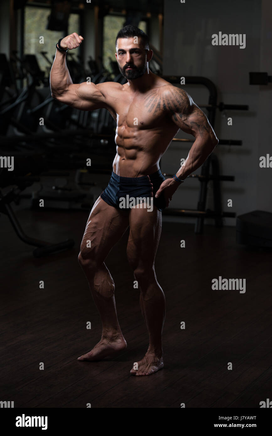 Beau jeune homme debout forte dans la salle de sport et Flexing Muscles ...