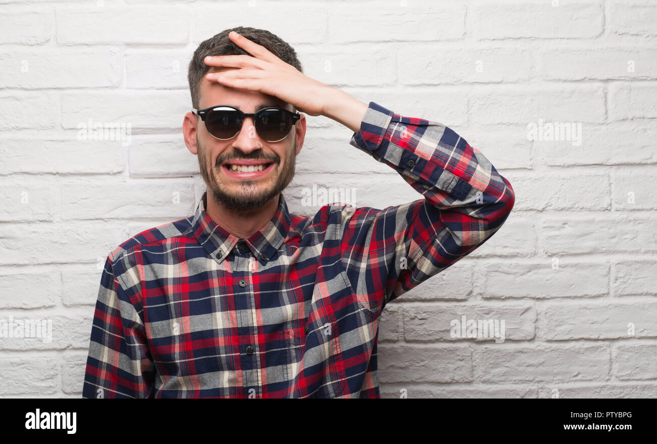 Young adult man wearing sunglasses standing over white brick wall a ...