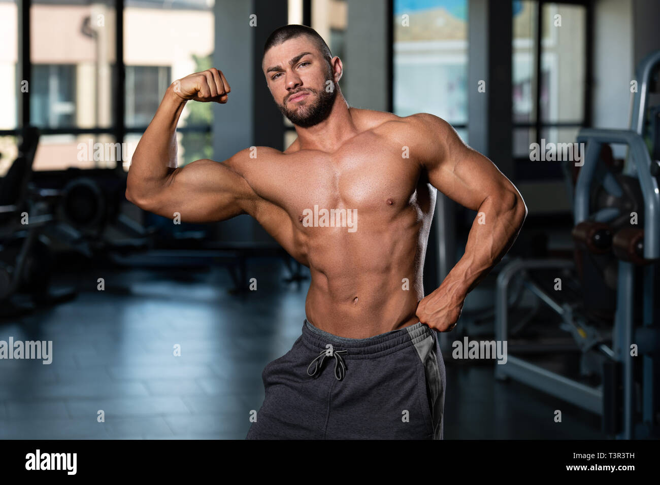 Beau jeune homme debout forte dans la salle de sport et Flexing Muscles ...