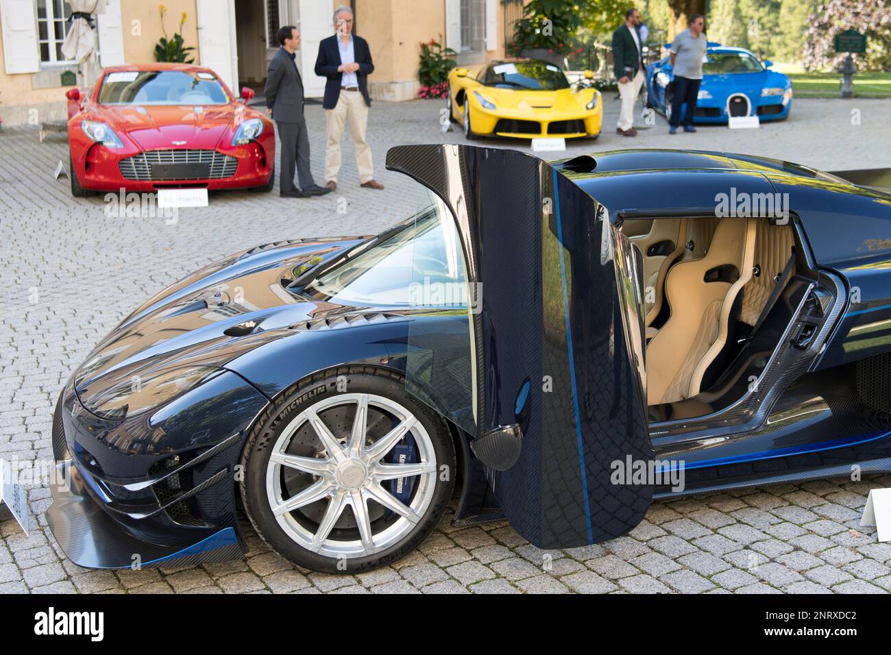 People looking at a 2015 Koenigsegg One:1 model car in front of a Aston ...