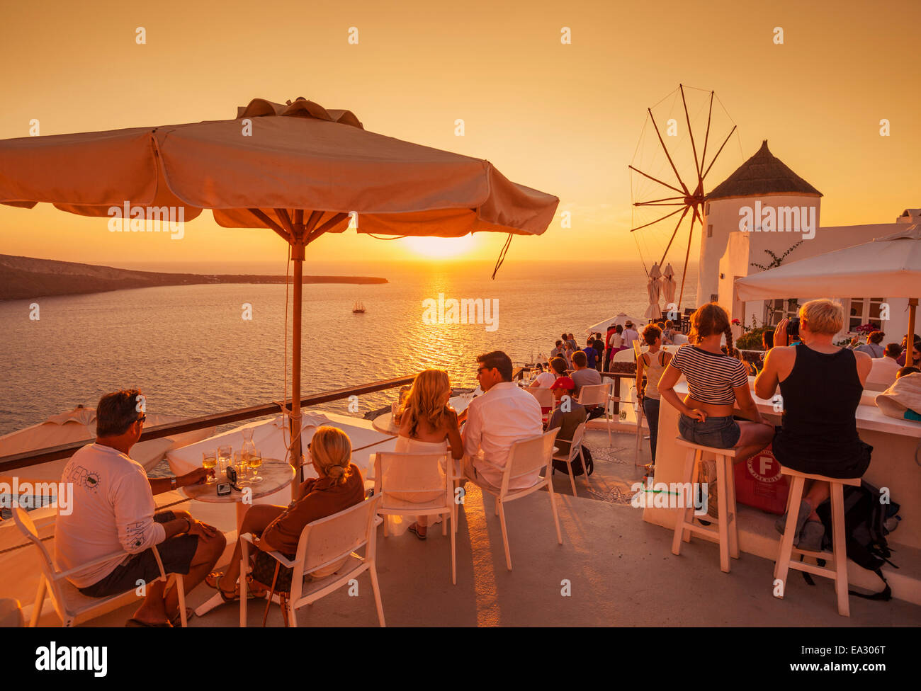 Bar affollato e mulino a vento al tramonto dal Mar Egeo, Oia ...