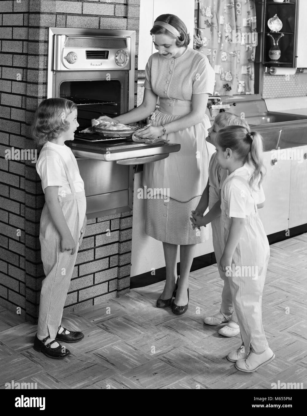 Anni Cinquanta madre e tre figlie in piedi intorno al forno in cucina ...